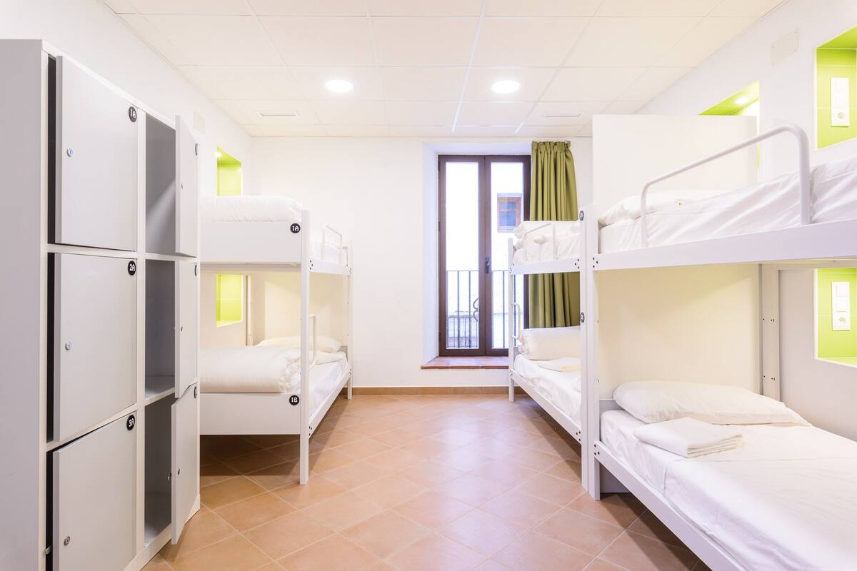 Um quarto de estudante com beliches, janela com cortinas e porta para uma varanda.