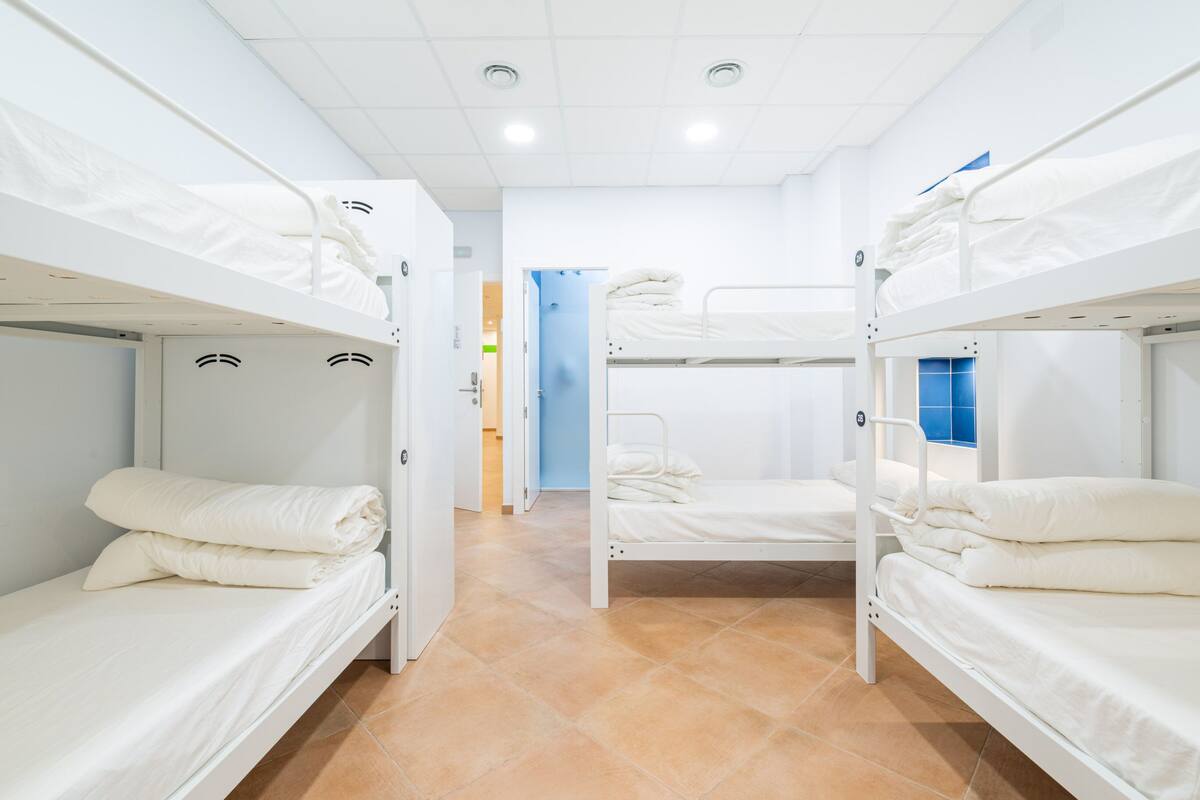 Quarto em dormitório limpo e bem iluminado, com beliches, piso ladrilhado e uma porta que dá para outro cômodo.