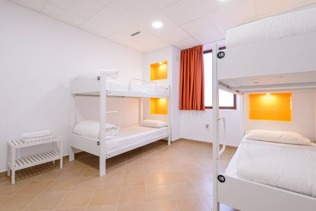 Quarto em dormitório com beliches, uma pequena mesa com uma toalha e uma janela com cortinas laranja.
