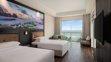 Quarto de hotel com duas camas, uma janela grande com vista para o oceano, uma televisão montada e uma pintura de uma paisagem costeira.