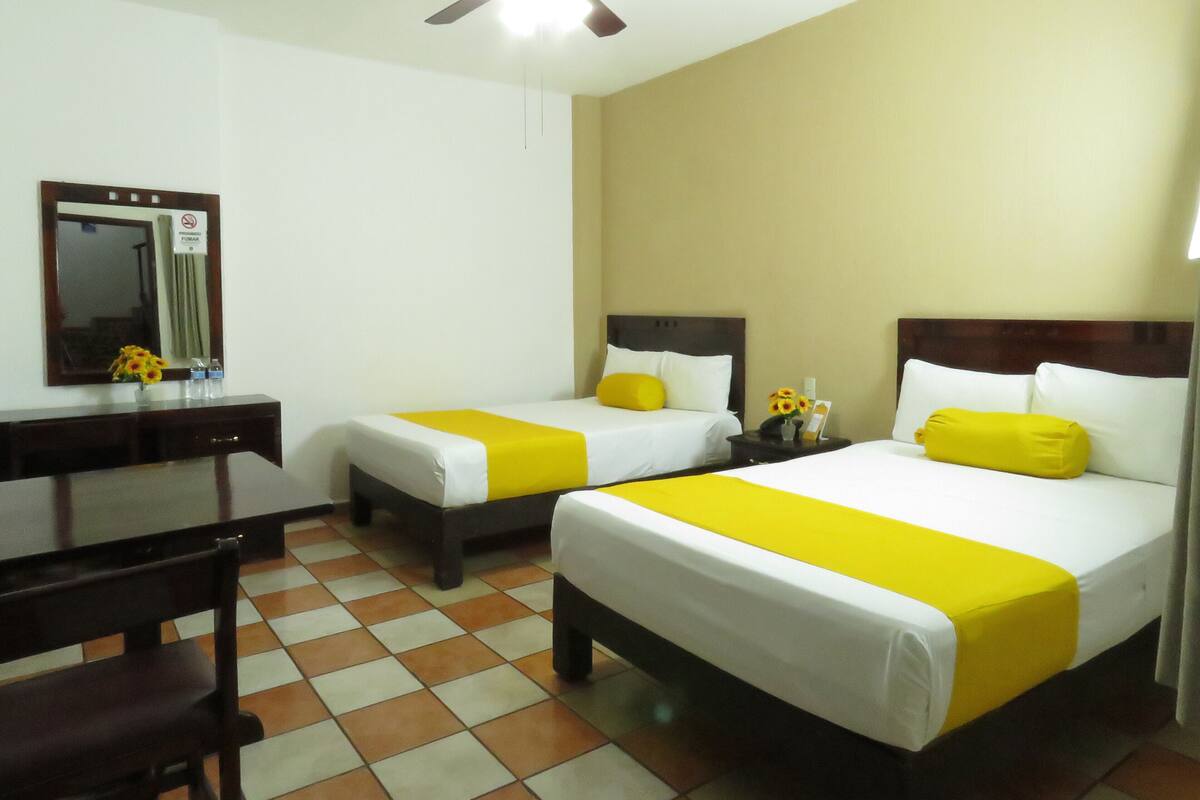 Quarto de hotel com duas camas, uma escrivaninha, uma cadeira, um ventilador de teto e um espelho.