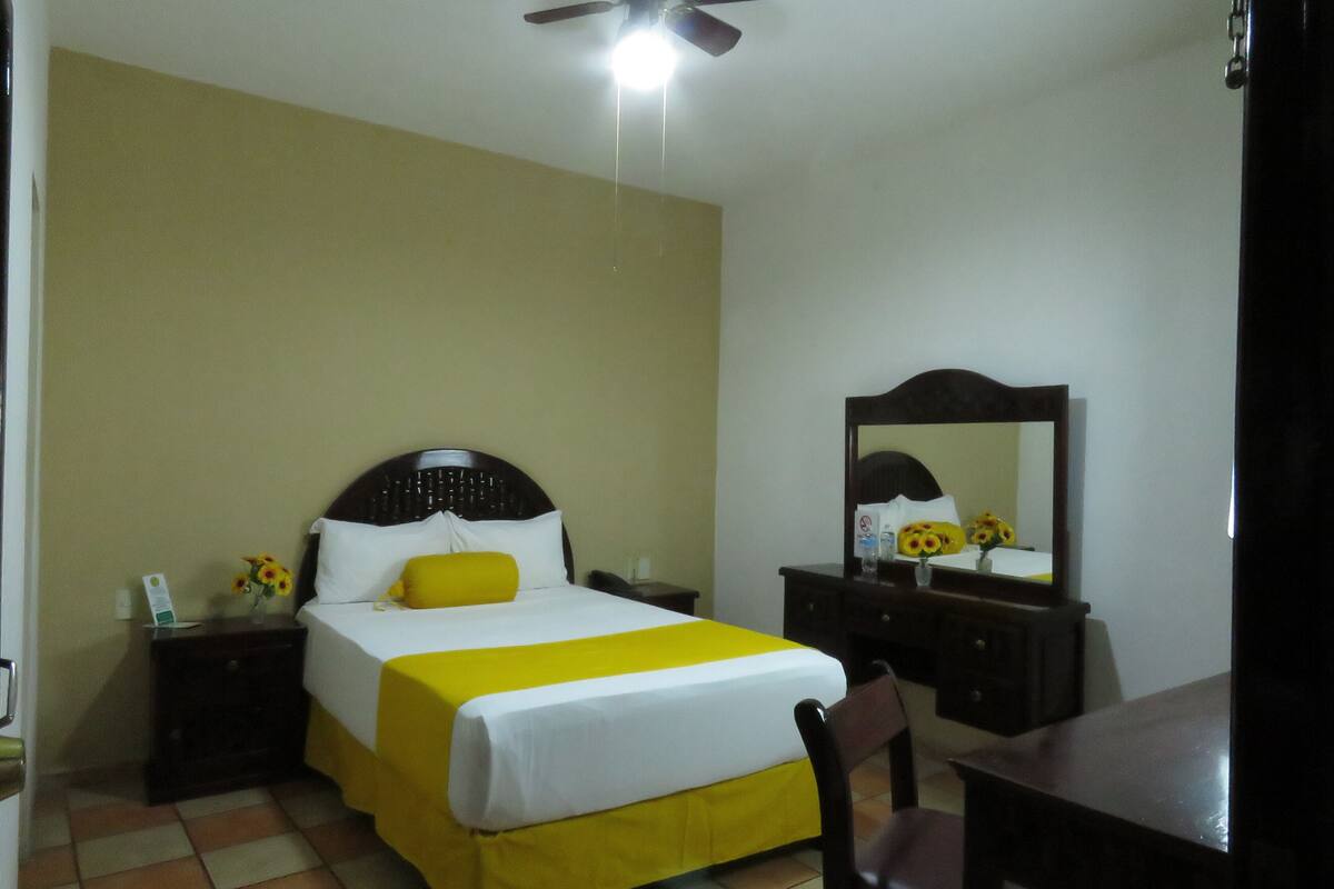 Quarto de hotel com cama, mesa de cabeceira, cadeira, mesa, espelho e ventilador de teto.