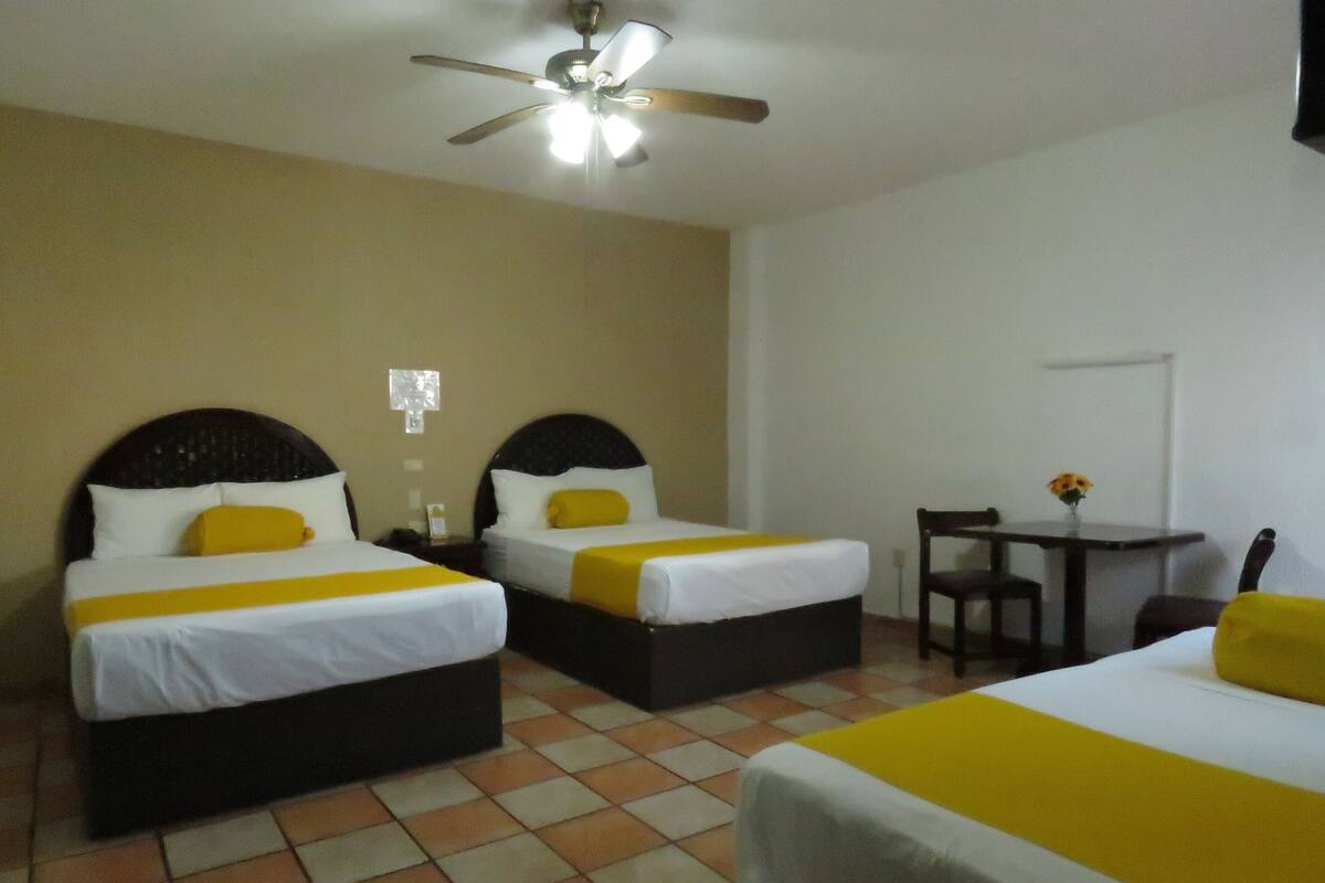 Quarto de hotel com duas camas, um ventilador de teto, uma mesa pequena e uma cadeira.