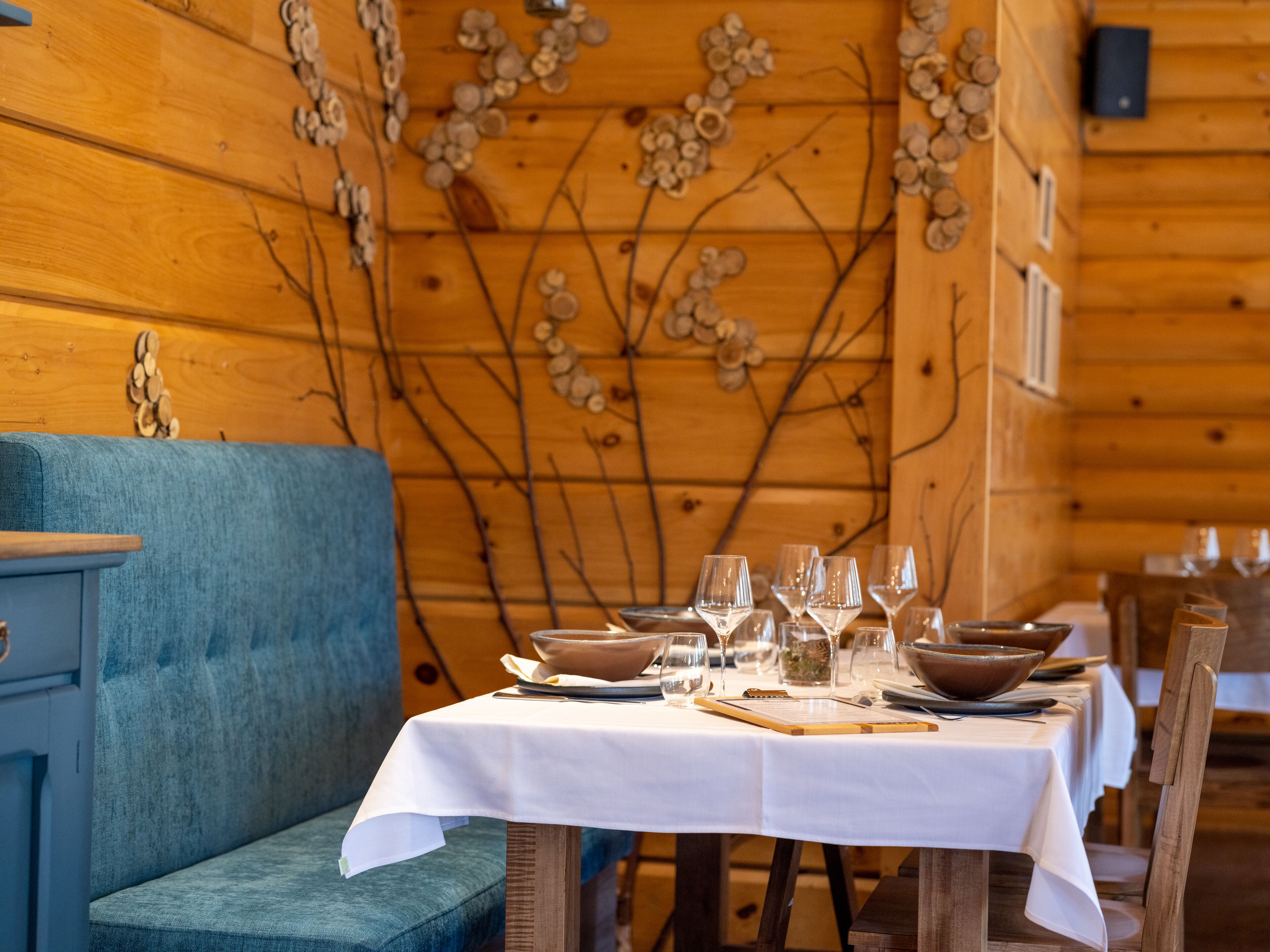 Interior de uma cabana de madeira com uma mesa de jantar posta para refeição, rodeada por paredes de madeira e um banco estofado azul.