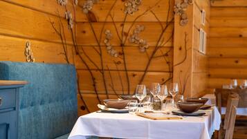 Interior de uma cabana de madeira com uma mesa de jantar posta para refeição, rodeada por paredes de madeira e um banco estofado azul.