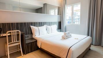 Quarto de hotel com cama, escrivaninha, cadeira e janela com cortinas.