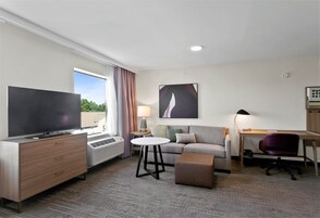 Quarto de hotel com televisão de tela plana em penteadeira de madeira, sofá com mesinha lateral, escrivaninha com cadeira e uma janela grande com cortinas.