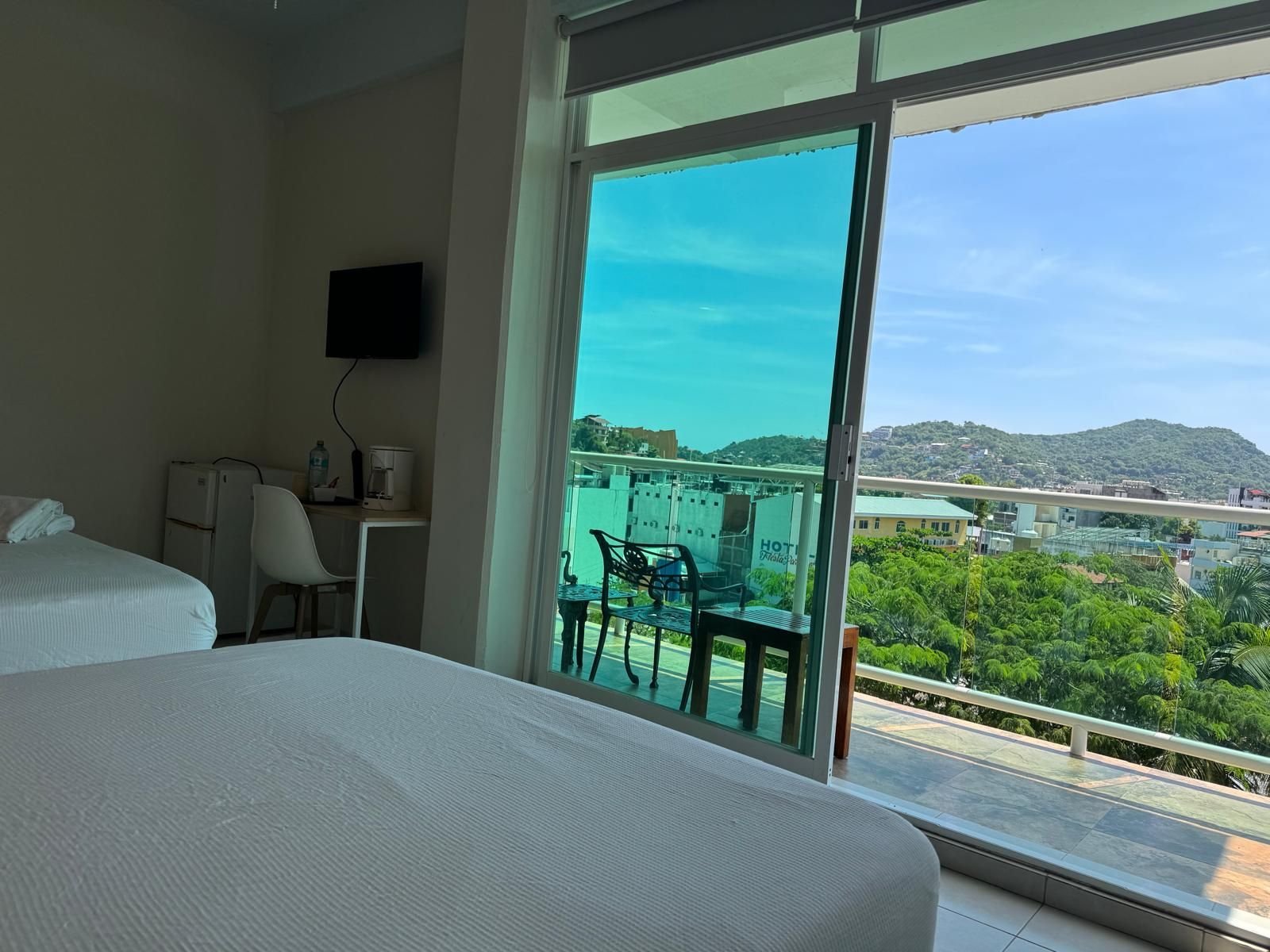 Quarto de hotel com cama, escrivaninha, cadeira, televisão e varanda com vista para a cidade e montanhas.