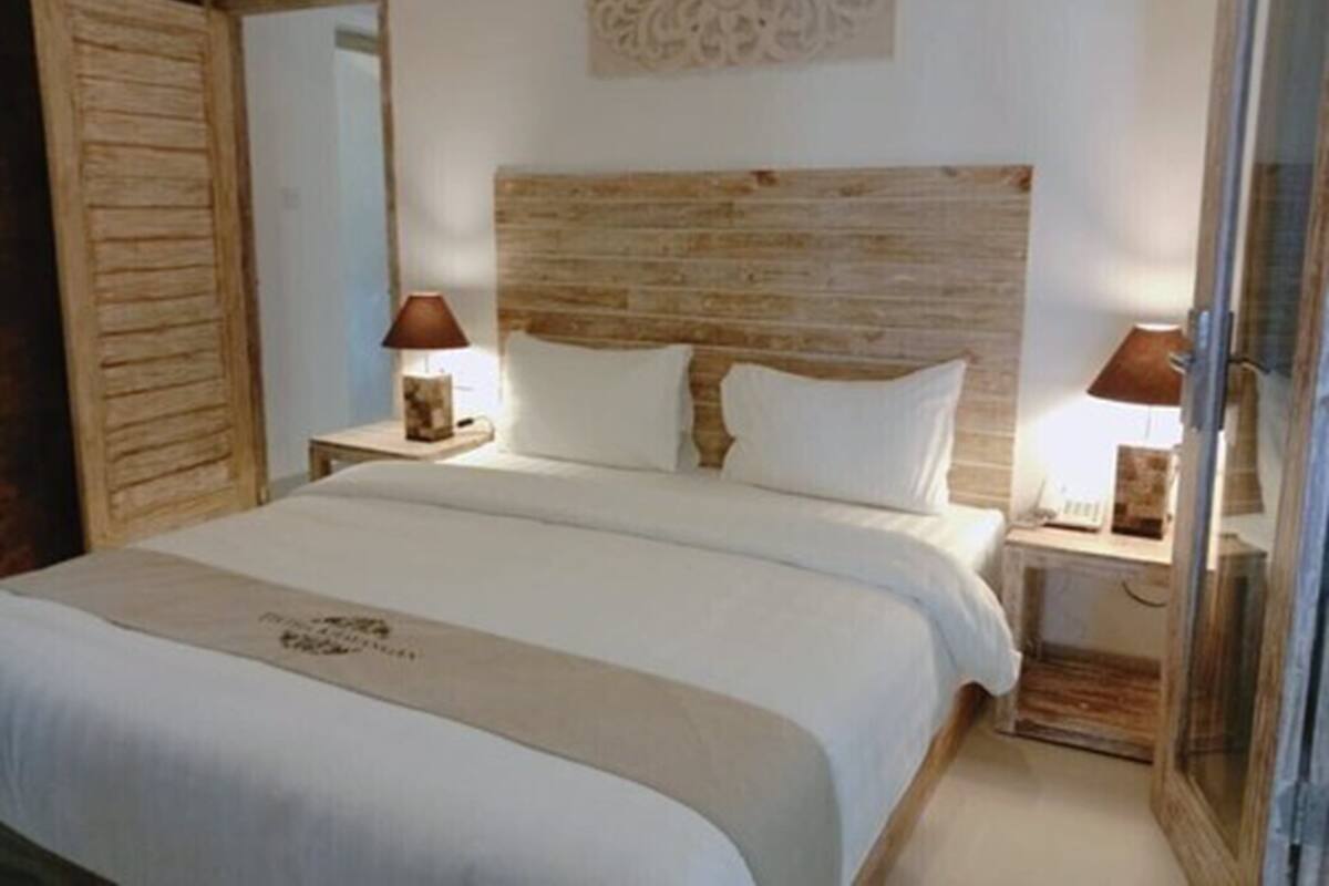 Quarto com cabeceira de madeira, cama com roupa de cama branca, duas mesas de cabeceira com abajures e um objeto decorativo na parede.