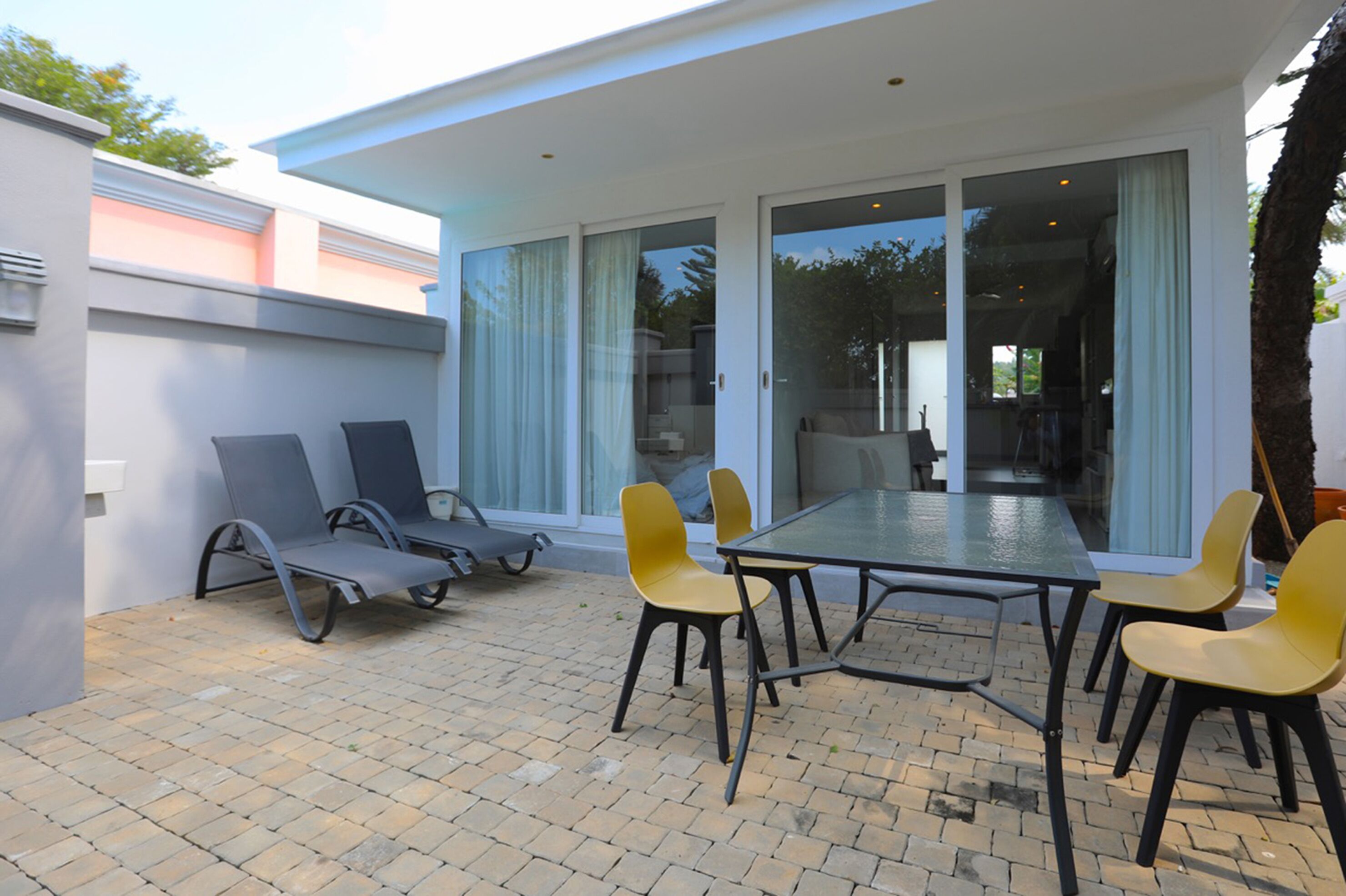 Um pátio externo com mesa e cadeiras de vidro, piso de tijolos e vista do interior através de grandes janelas.