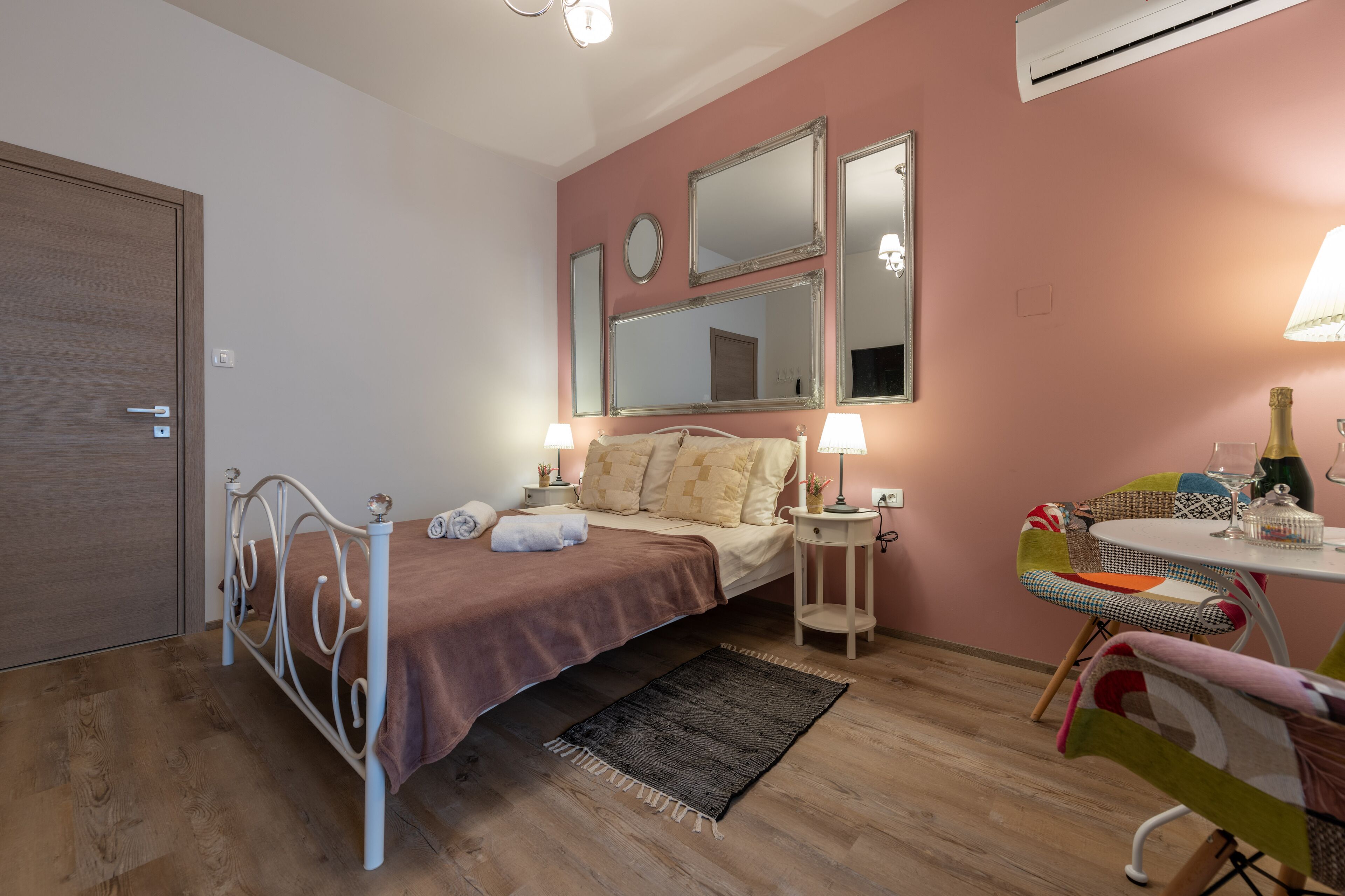 Quarto com cama, mesas de cabeceira, espelho, cadeira e uma pequena mesa com abajur.