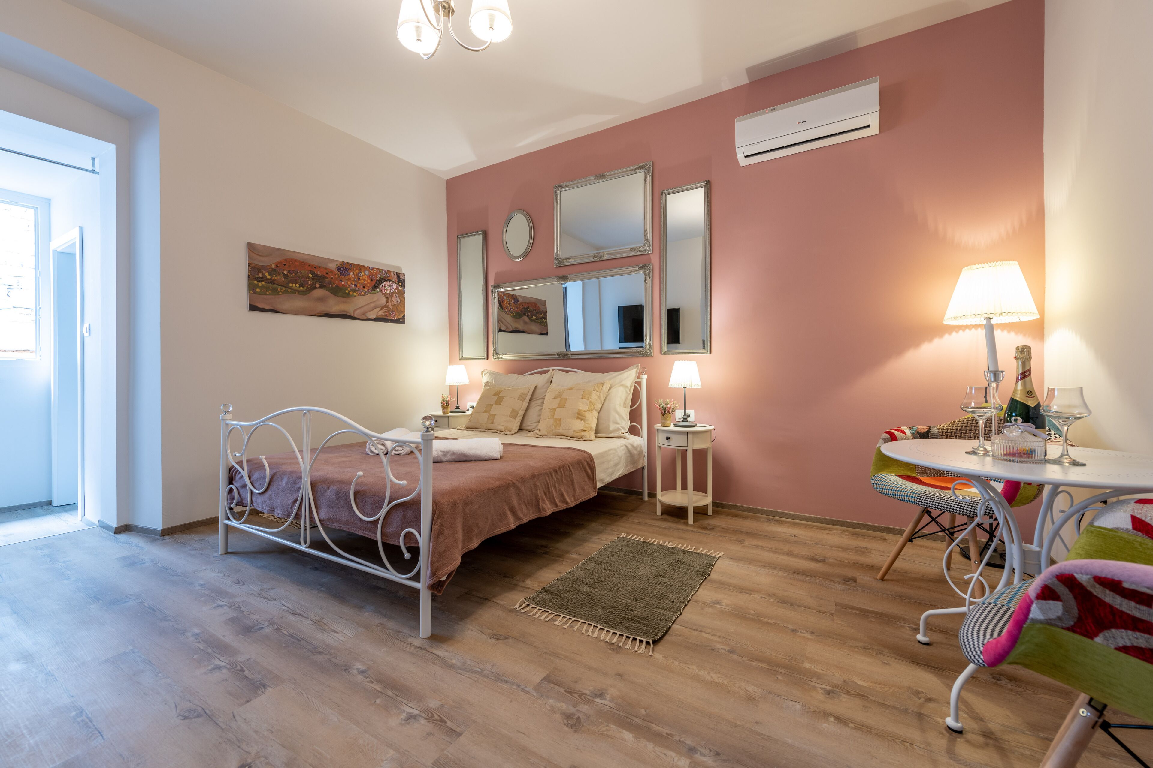 Quarto com estrutura de cama de metal, mesa de cabeceira com abajur, mesinha redonda com abajur, uma cadeira e um tapete.