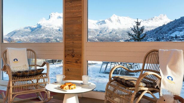 Quarto aconchegante com duas cadeiras de vime, uma mesa pequena com doces e vista para montanhas cobertas de neve.