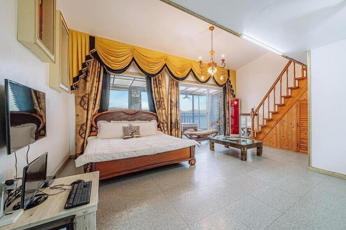 Quarto com cama, escrivaninha com computador, cadeira, mesa, escada e janela com cortinas.