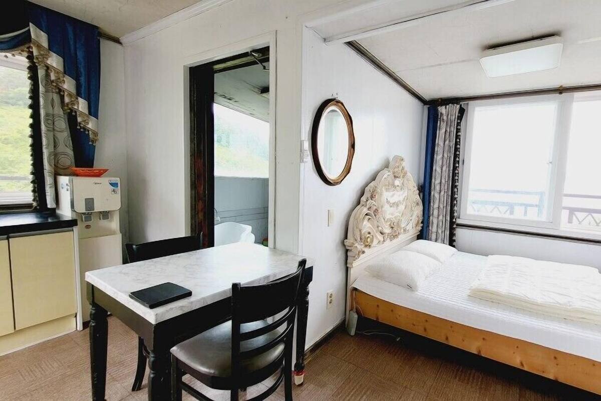 Quarto pequeno e compacto com uma cama de solteiro, uma mesa e uma cadeira. Há uma janela que oferece luz natural e vista para o exterior.