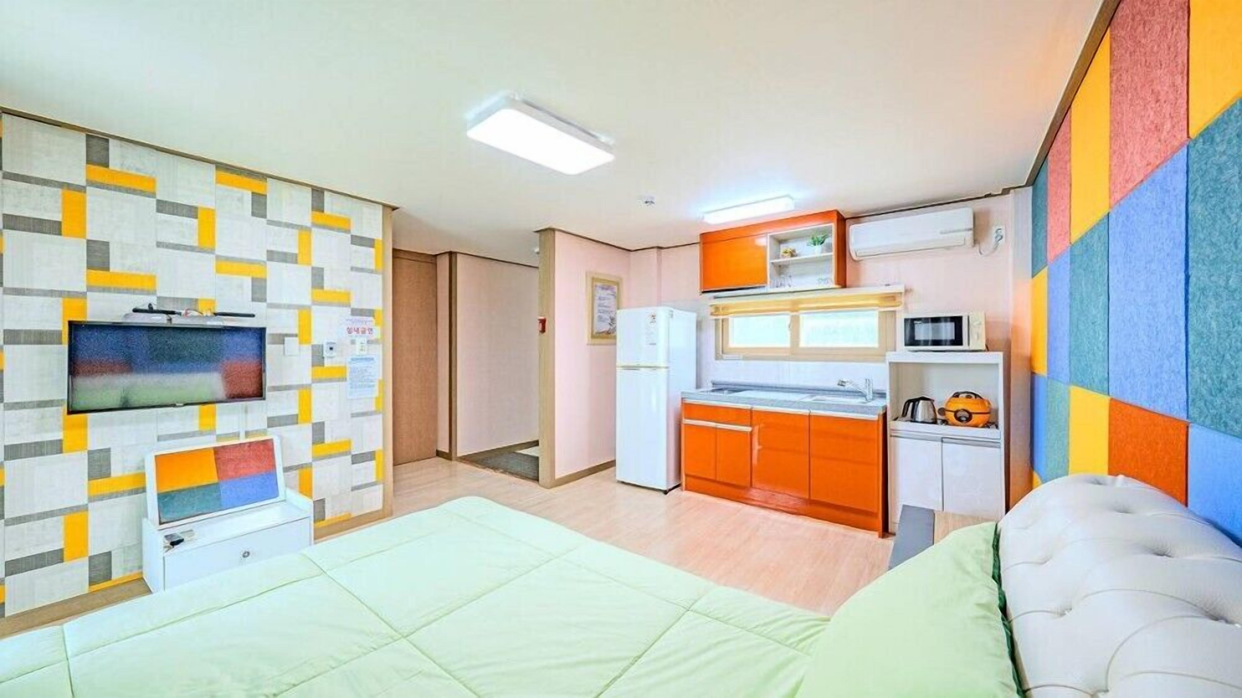 Quarto de hotel compacto com cama, pequena cozinha, televisão e parede colorida.