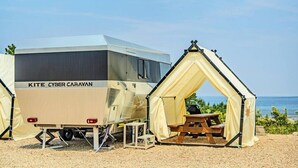 Uma caravana moderna com uma tenda acoplada, incluindo uma mesa e cadeiras, localizada na praia, com o mar ao fundo.