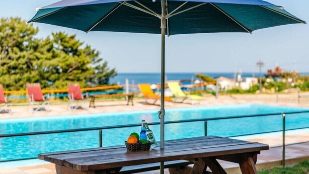 Área da piscina com mesa de madeira, guarda-sol azul e vista para o oceano.