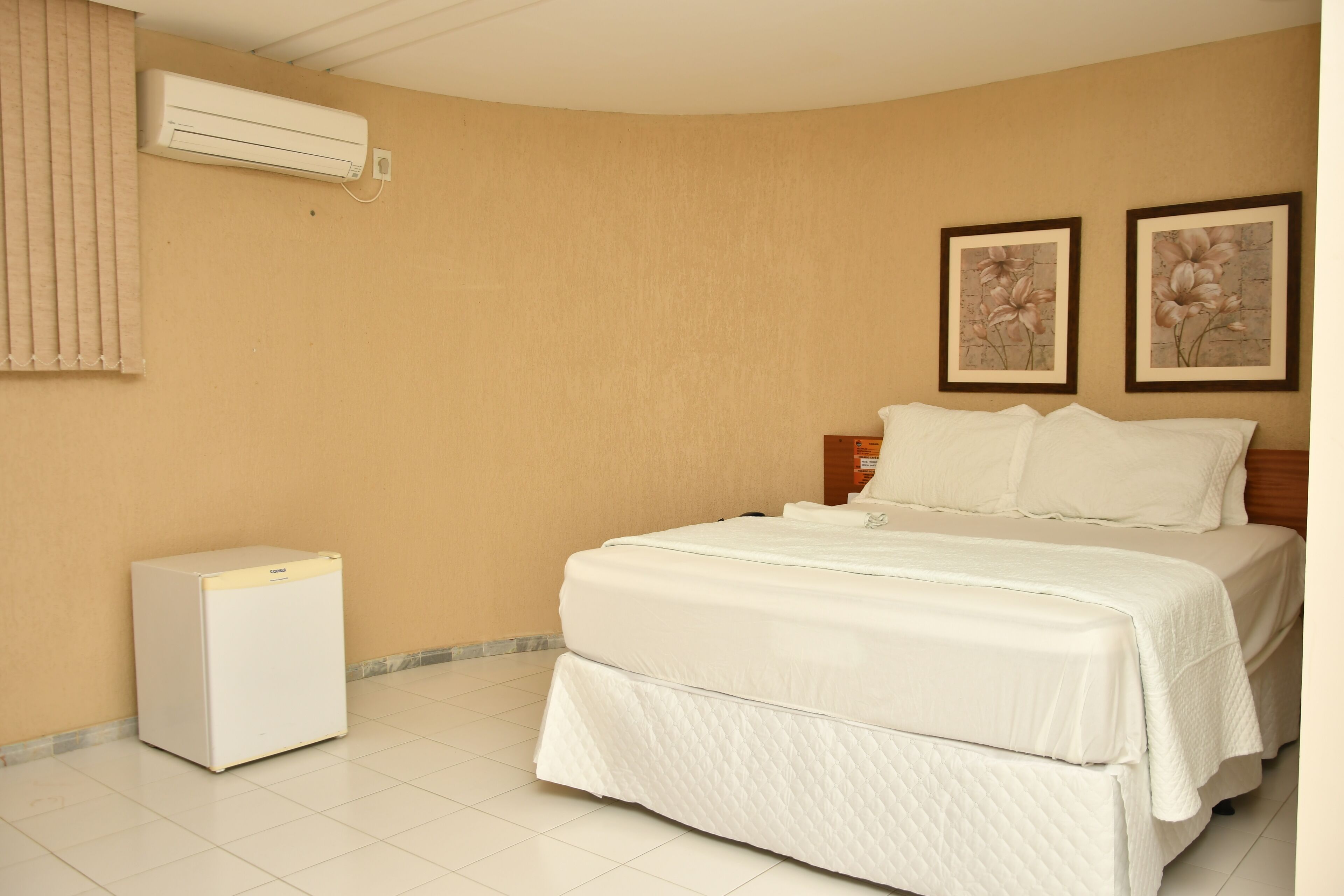 Quarto de hotel com uma cama, um pequeno frigorífico branco e duas imagens emolduradas com flores na parede.