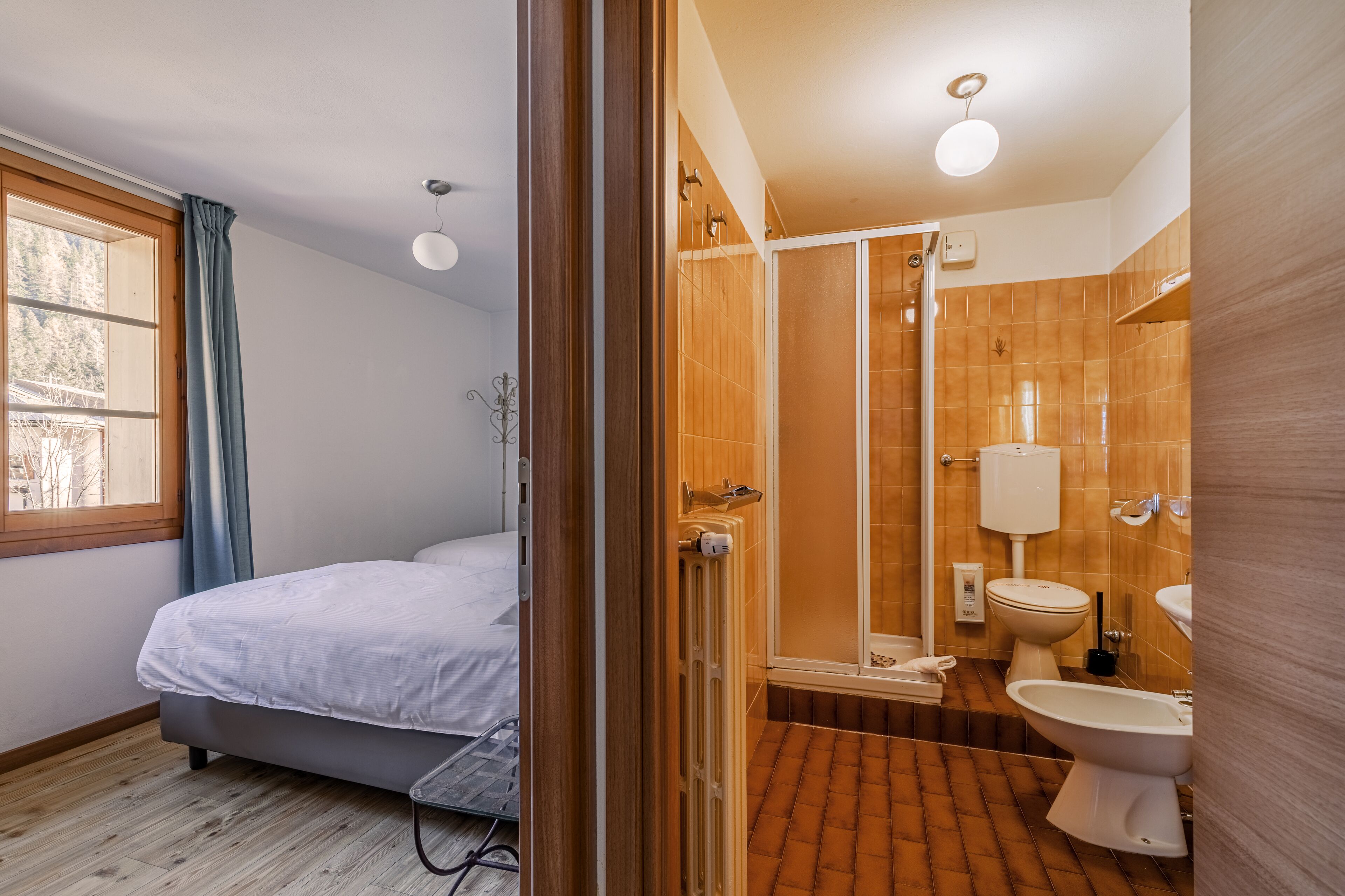 Quarto com cama, janela com cortinas e chão de madeira. O banheiro possui chuveiro, vaso sanitário e bidê.