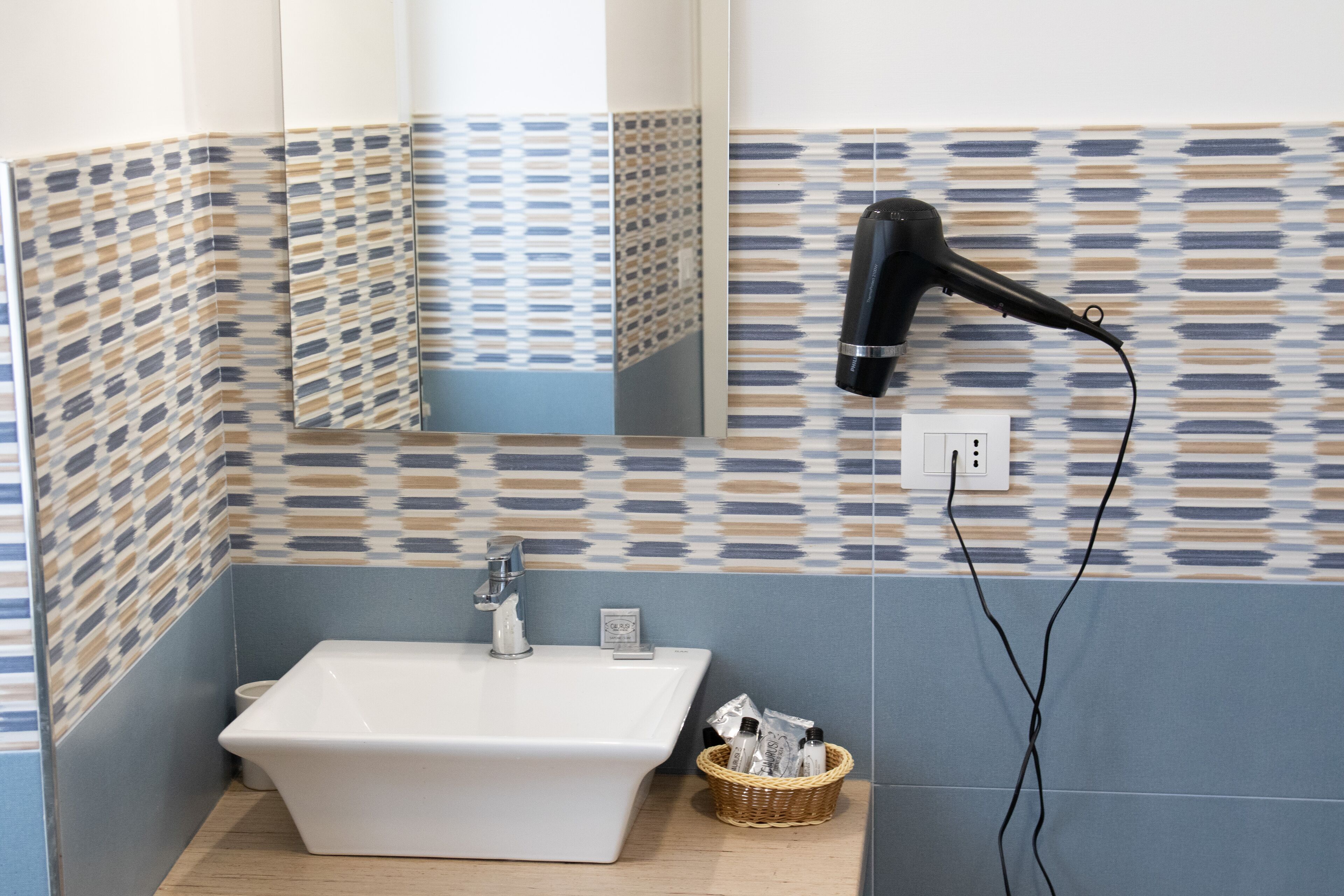 Casa de banho com lavatório branco, espelho, secador de cabelo e parede de azulejos estampados.