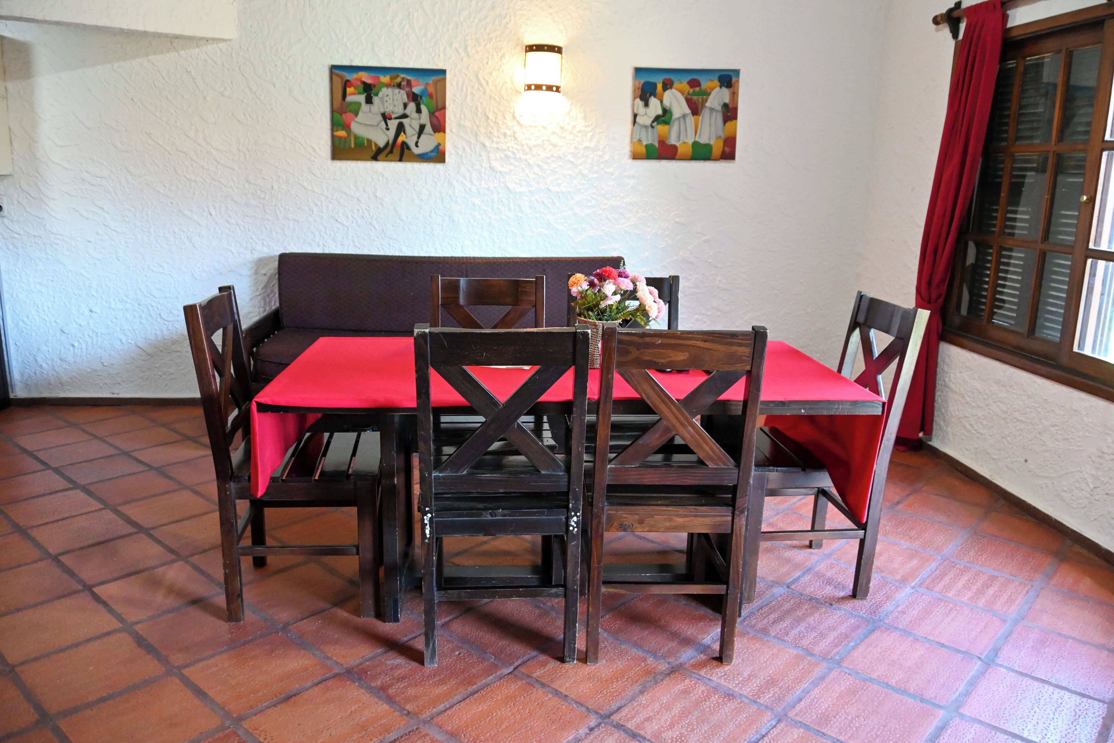 Uma sala de jantar com uma mesa vermelha e quatro cadeiras de madeira, uma parede branca com textura e duas luminárias fixadas na parede.