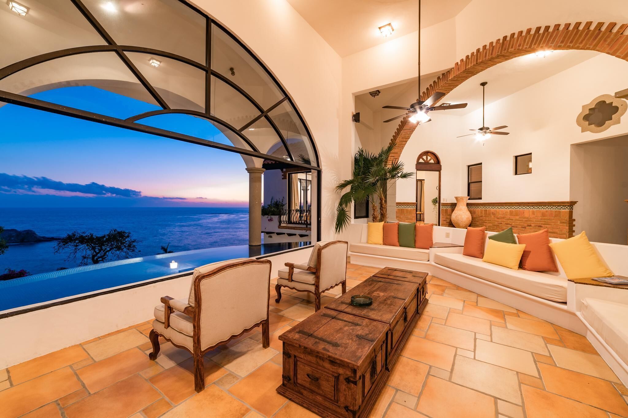 Ampla sala de estar com uma grande janela em arco com vista para uma piscina e o oceano, um espaço de estar acolhedor com almofadas e um ventilador de teto.