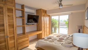 Quarto com cama, televisão em prateleira de madeira, ventilador de teto e vista para o verde lá fora.