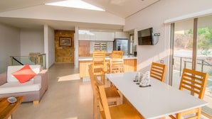 Uma sala de estar moderna com sofá, mesa de jantar e cadeiras. Há uma área de cozinha com armários e geladeira. É visível um ventilador de teto, e há uma TV fixada na parede.