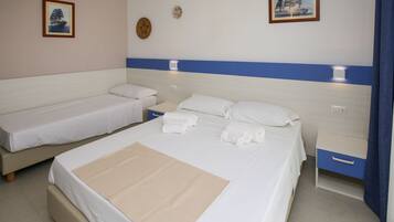 Quarto de hotel com duas camas de solteiro, uma cama maior com manta bege, mesa de cabeceira com luminária e um pequeno móvel com gavetas.