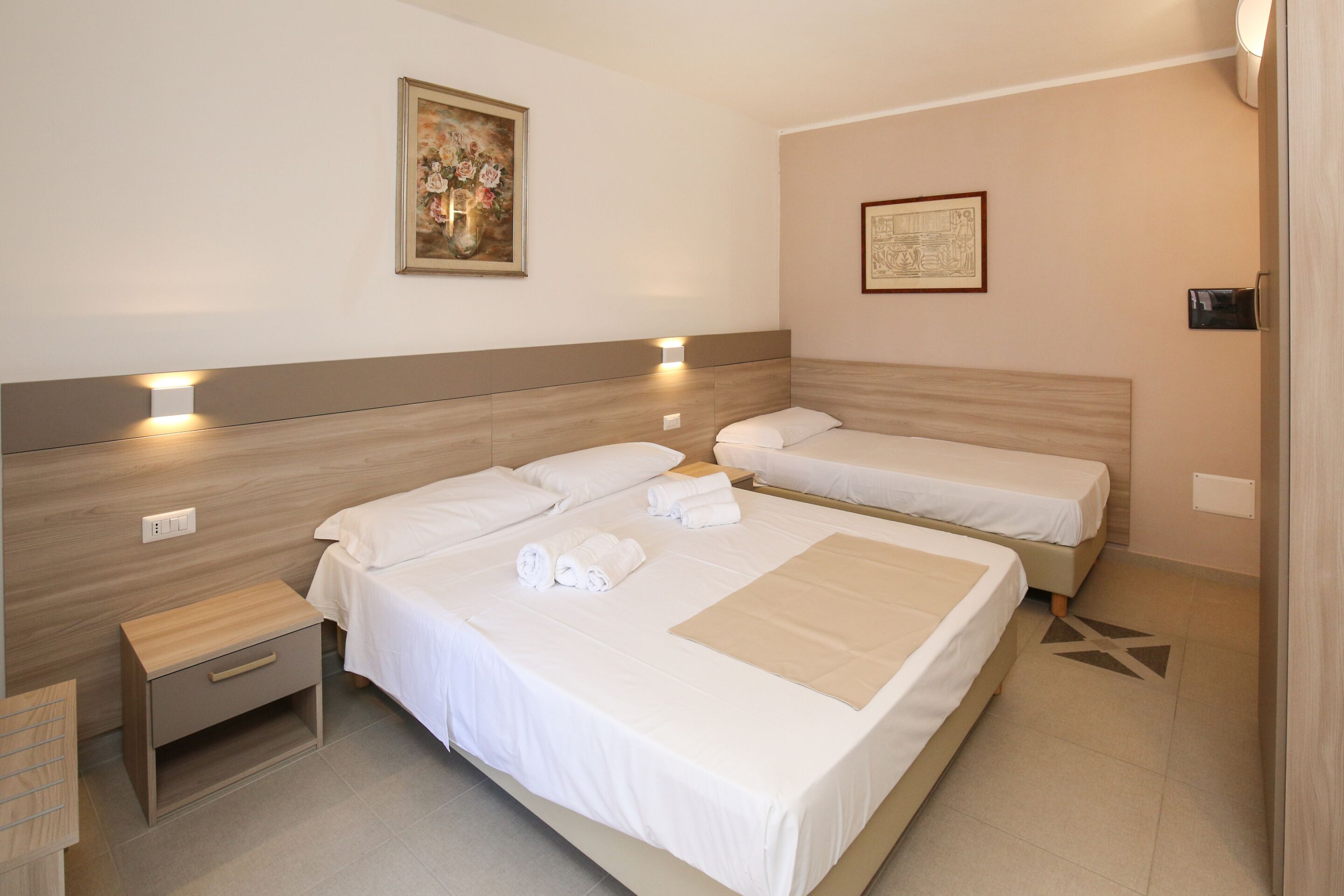 Quarto de hotel com duas camas, mesa de cabeceira e luminárias na parede.