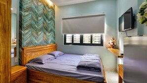Quarto com cama, cabeceira de madeira, janela com persianas, televisão na parede e mesa de cabeceira de madeira.