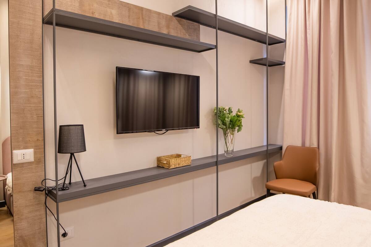Quarto moderno com uma cama grande, televisão na parede, mesa pequena com luminária e uma cadeira.