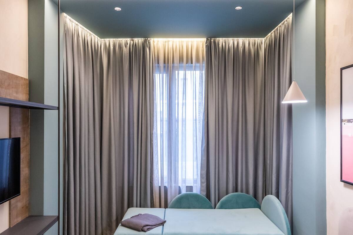 Quarto de hotel moderno com cama, televisão, mesa pequena e janela com cortinas.