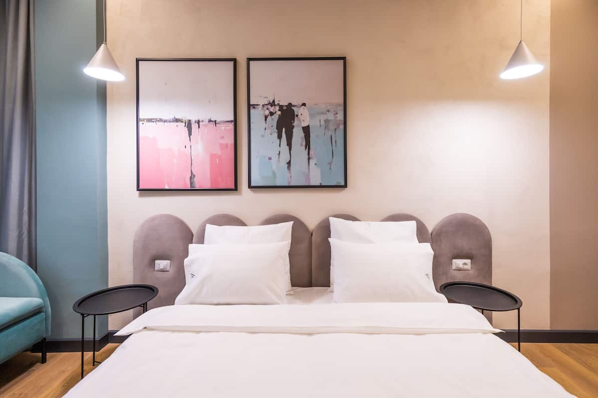Quarto de hotel com cama, duas mesas de cabeceira, uma cadeira e duas pinturas penduradas na parede.