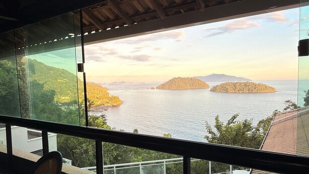 Quarto com uma grande janela com vista para uma paisagem costeira, com colinas e um corpo d'água.