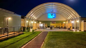 Uma área externa ampla e coberta, com um espaço de convivência central e uma tela de televisão.