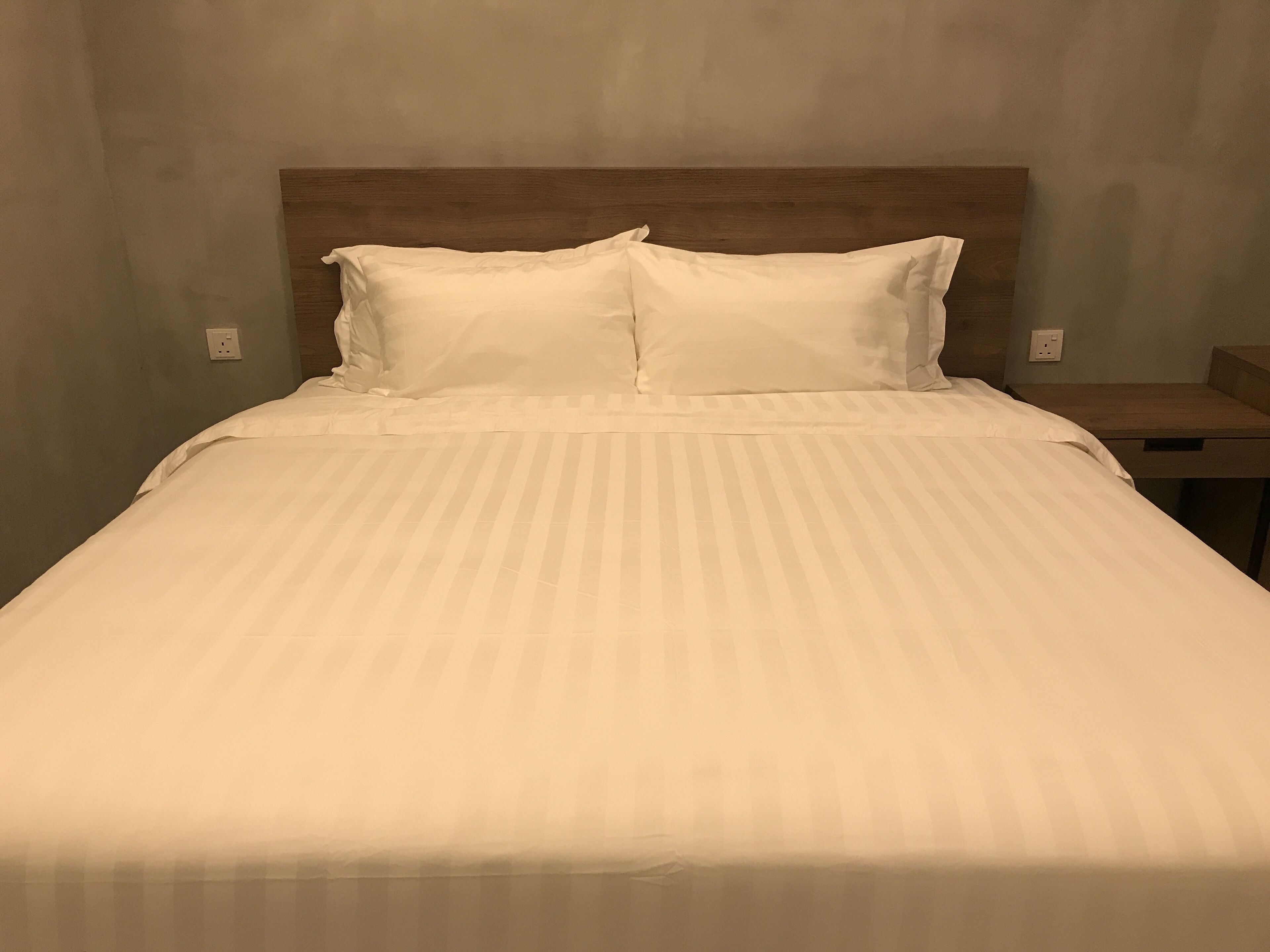 Cama bem arrumada com cabeceira de madeira, parede lisa e mesa de cabeceira pequena com tomada.