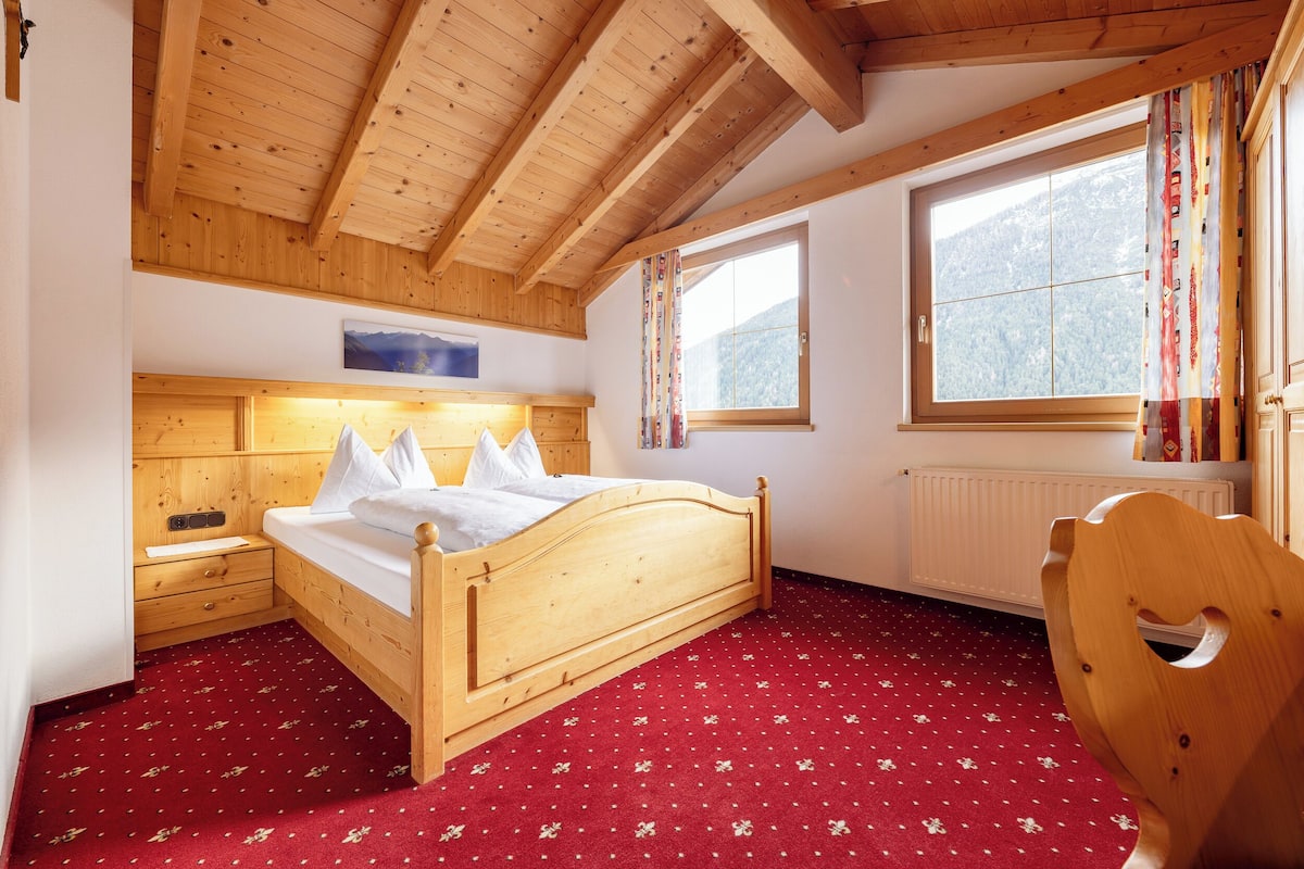 Quarto de madeira com uma cama, uma mesa de cabeceira, uma cadeira e uma janela com vista para montanhas.