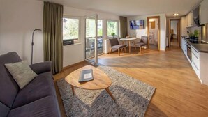 Sala de estar moderna com um sofá escuro, mesa de centro de madeira e tapete cinza. Há um espaço de refeições com mesa e cadeiras, e uma cozinha com armários e uma TV.