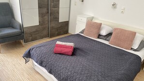 Uma cama bem arrumada com um edredom escuro estampado e duas almofadas, uma poltrona cinza, uma mesa de cabeceira pequena com abajur e um guarda-roupa com espelho.