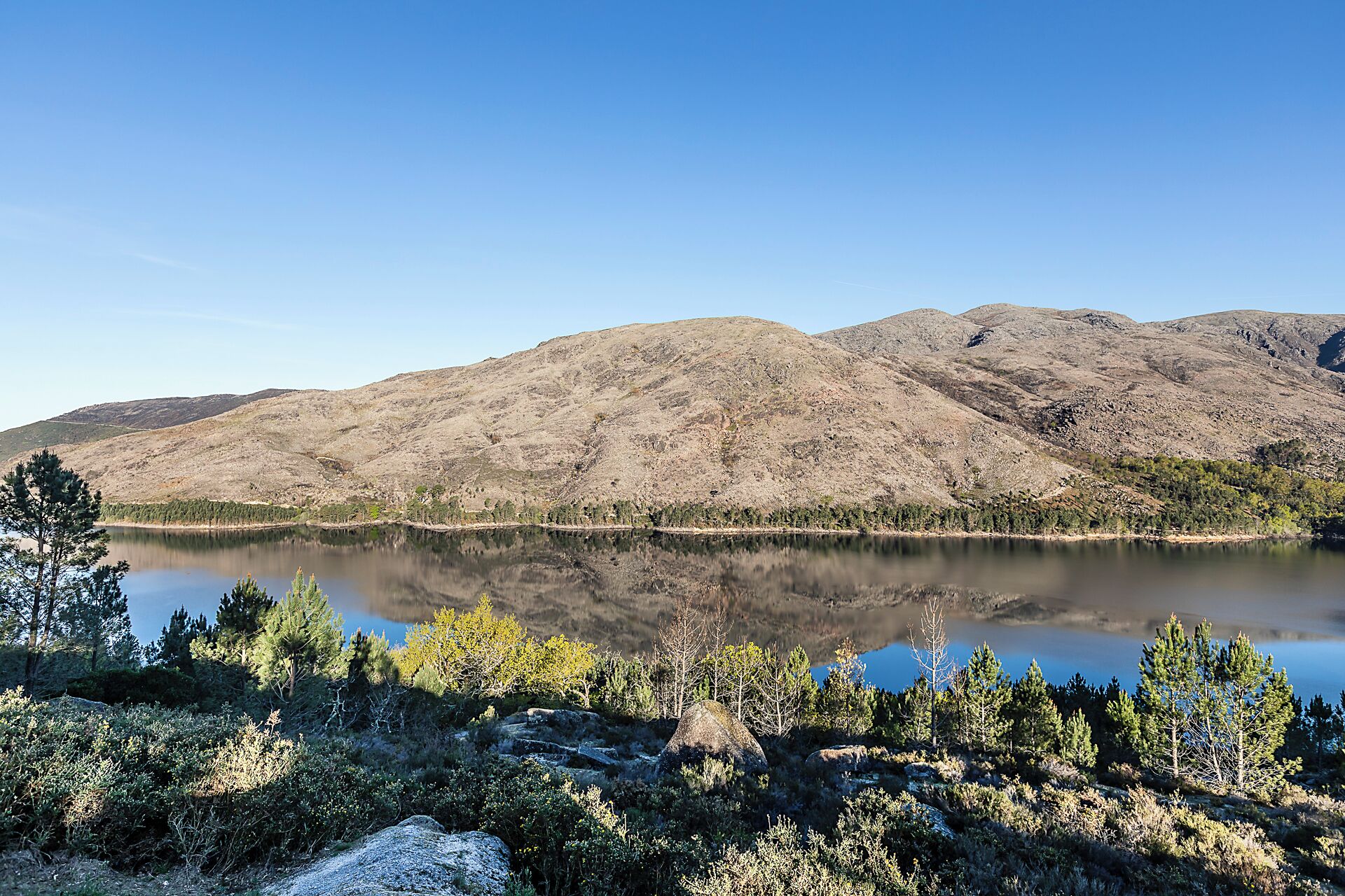 Paisagem montanhosa com um lago tranquilo, rodeado por floresta densa e terreno rochoso.