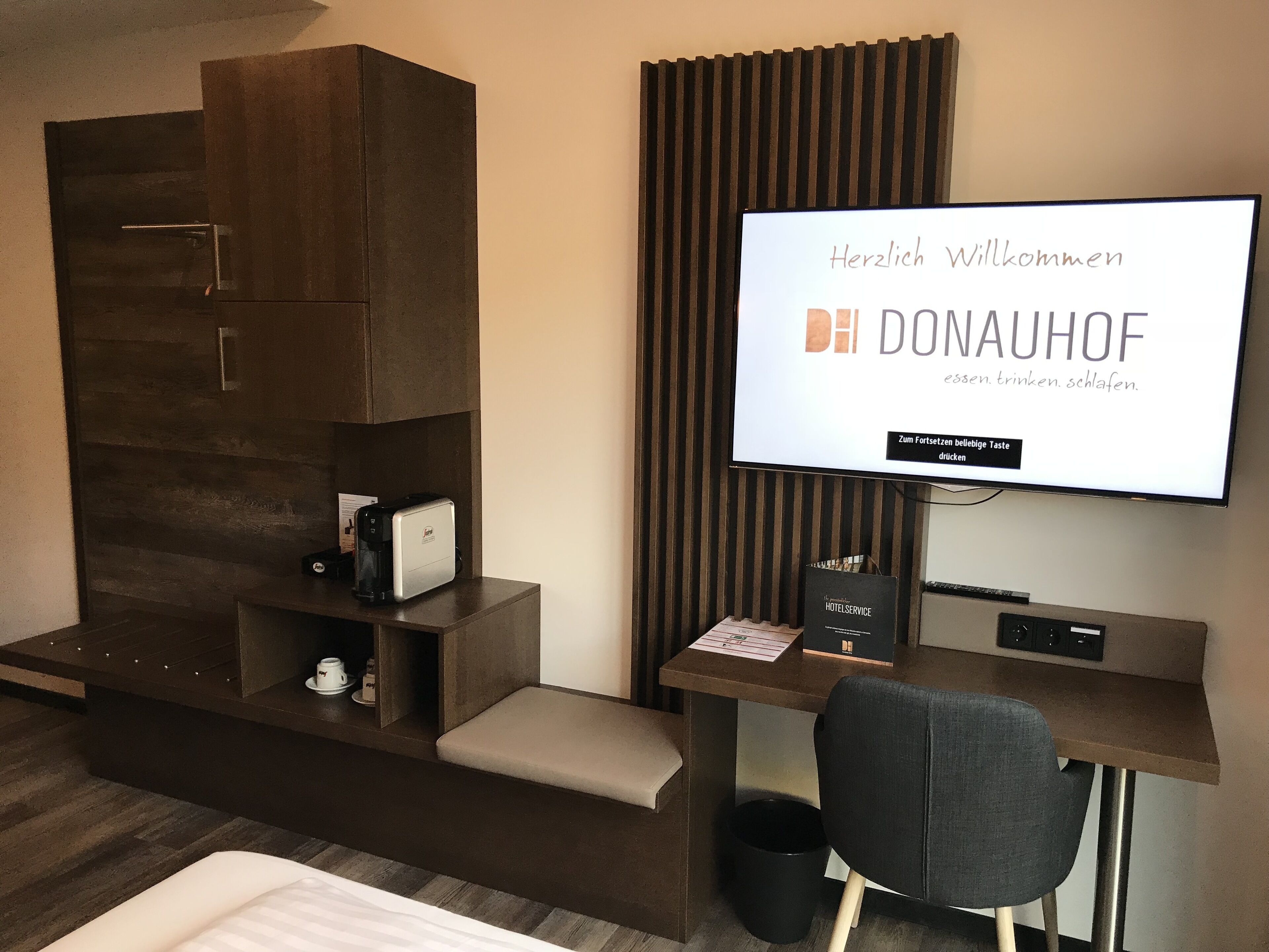 Quarto de hotel moderno com uma televisão de tela plana grande exibindo uma mensagem de boas-vindas, uma máquina de café, uma escrivaninha com cadeira e um guarda-roupa embutido.