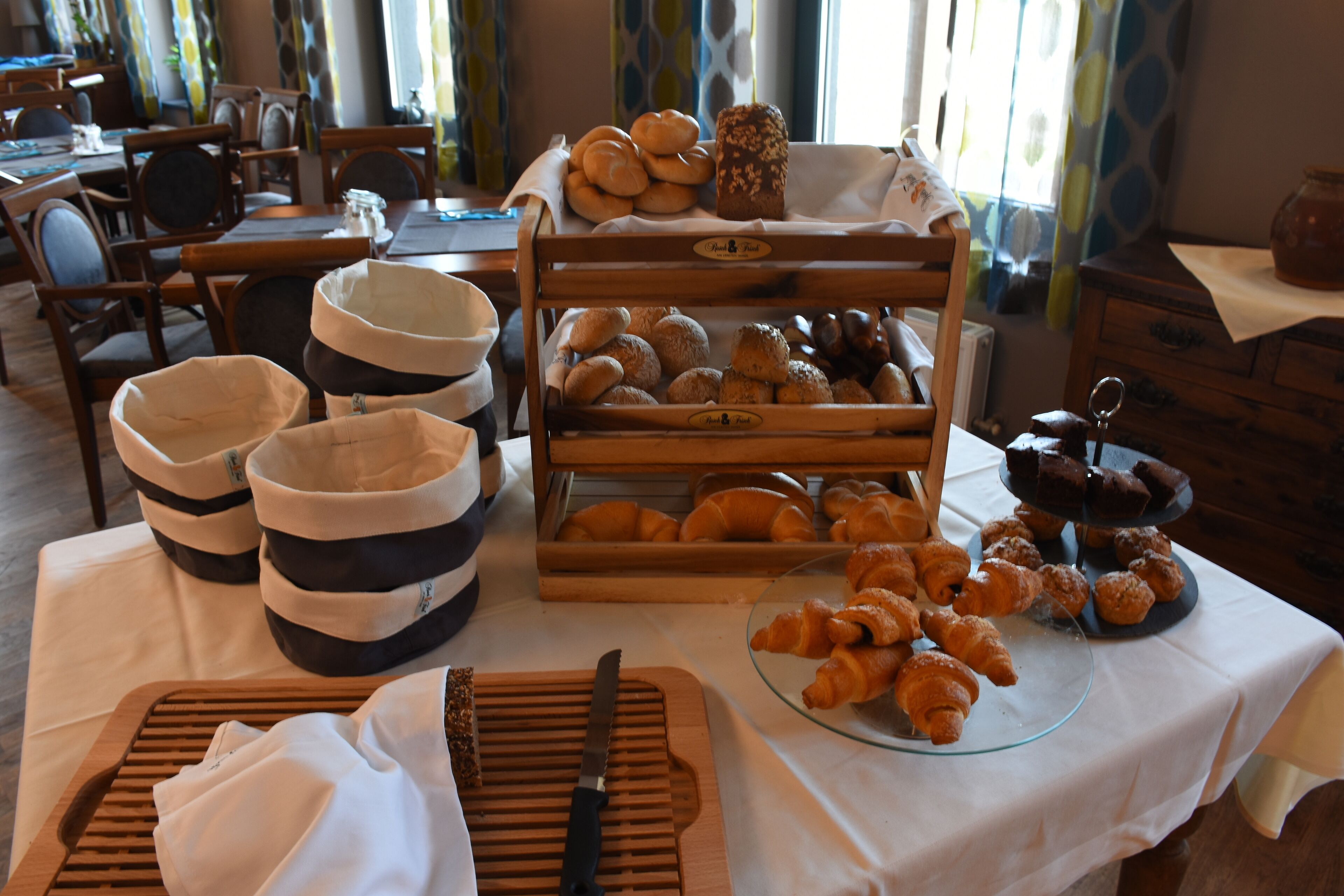 Buffet de café da manhã com diversos pães e doces, tábua de corte com faca e um recipiente com croissants.