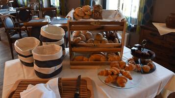 Buffet de café da manhã com diversos pães e doces, tábua de corte com faca e um recipiente com croissants.