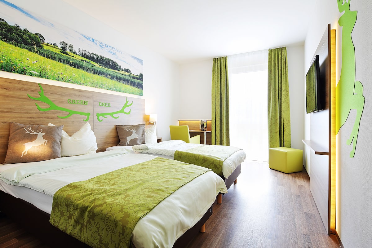 Quarto de hotel com duas camas, uma escrivaninha e decoração com tons de verde.