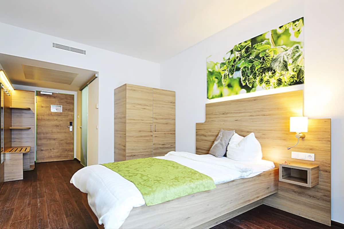 Quarto de hotel moderno com cama de madeira, manta verde, mesa de cabeceira e luminária fixada na parede.