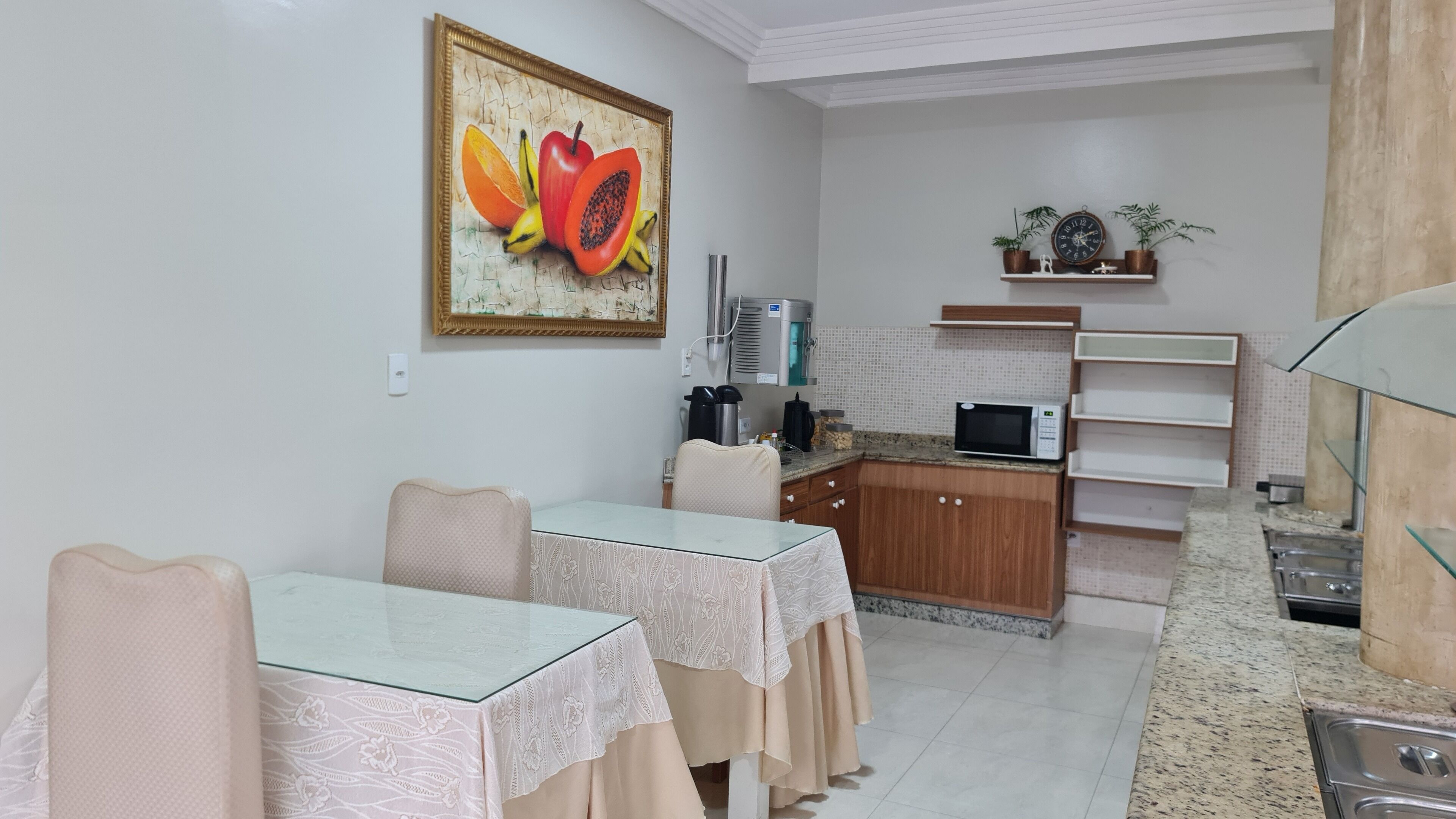 Uma sala de jantar com duas mesas de vidro, cadeiras bege, uma cozinha com micro-ondas e um relógio fixado na parede.