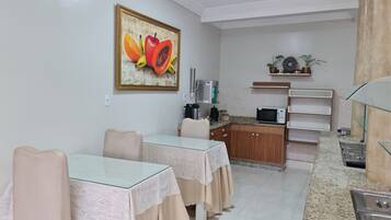 Uma sala de jantar com duas mesas de vidro, cadeiras bege, uma cozinha com micro-ondas e um relógio fixado na parede.