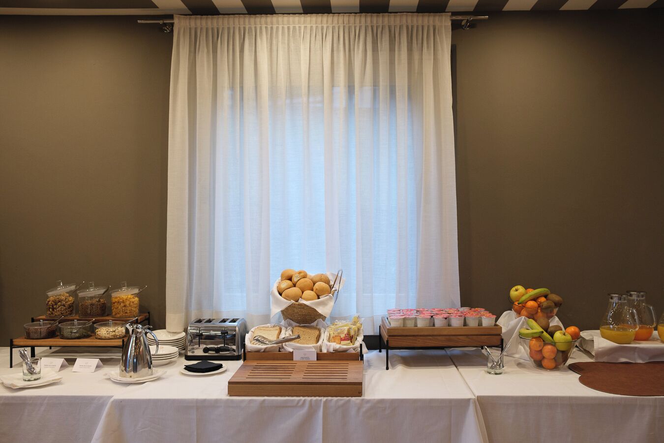 Disposição de buffet de café da manhã com diversos alimentos, incluindo pães, doces e frutas, sobre uma mesa com toalha branca.